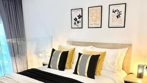 Quarto moderno com uma cama grande, cabeceira de madeira e uma mesa de cabeceira com abajur. Três obras de arte emolduradas estão na parede acima da cama.