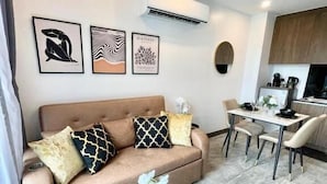 Uma sala de estar moderna com um sofá bege, uma mesa de jantar com cadeiras e uma cozinha ao fundo.