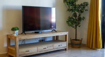 Uma TV de tela plana sobre um móvel de madeira com gavetas, uma pequena planta em vaso e uma árvore de interior alta.