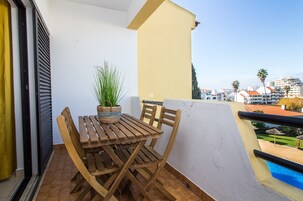 Uma varanda com mesa e cadeiras de madeira, um vaso com planta e vista para edifícios e palmeiras.