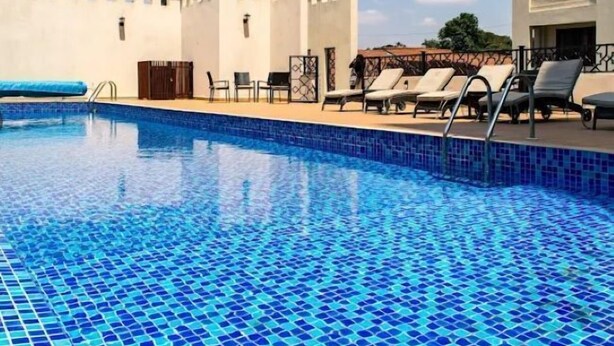Piscina com azulejos de mosaico azul, rodeada por cadeiras de espreguiçadeira e um edifício branco com fachada semelhante a um castelo.