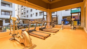 Ginásio com esteiras, elípticos e bicicleta ergométrica, com vista para uma área residencial através de amplas janelas.