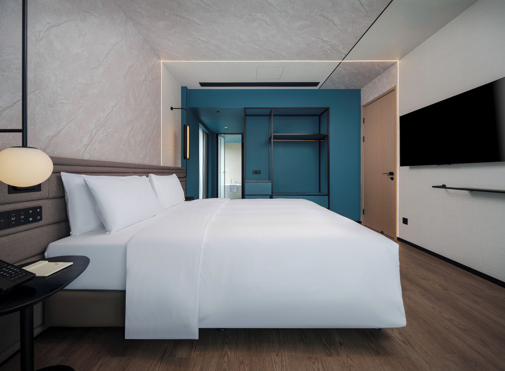 Quarto de hotel moderno com uma cama grande, uma escrivaninha com luminária e uma TV fixada na parede.