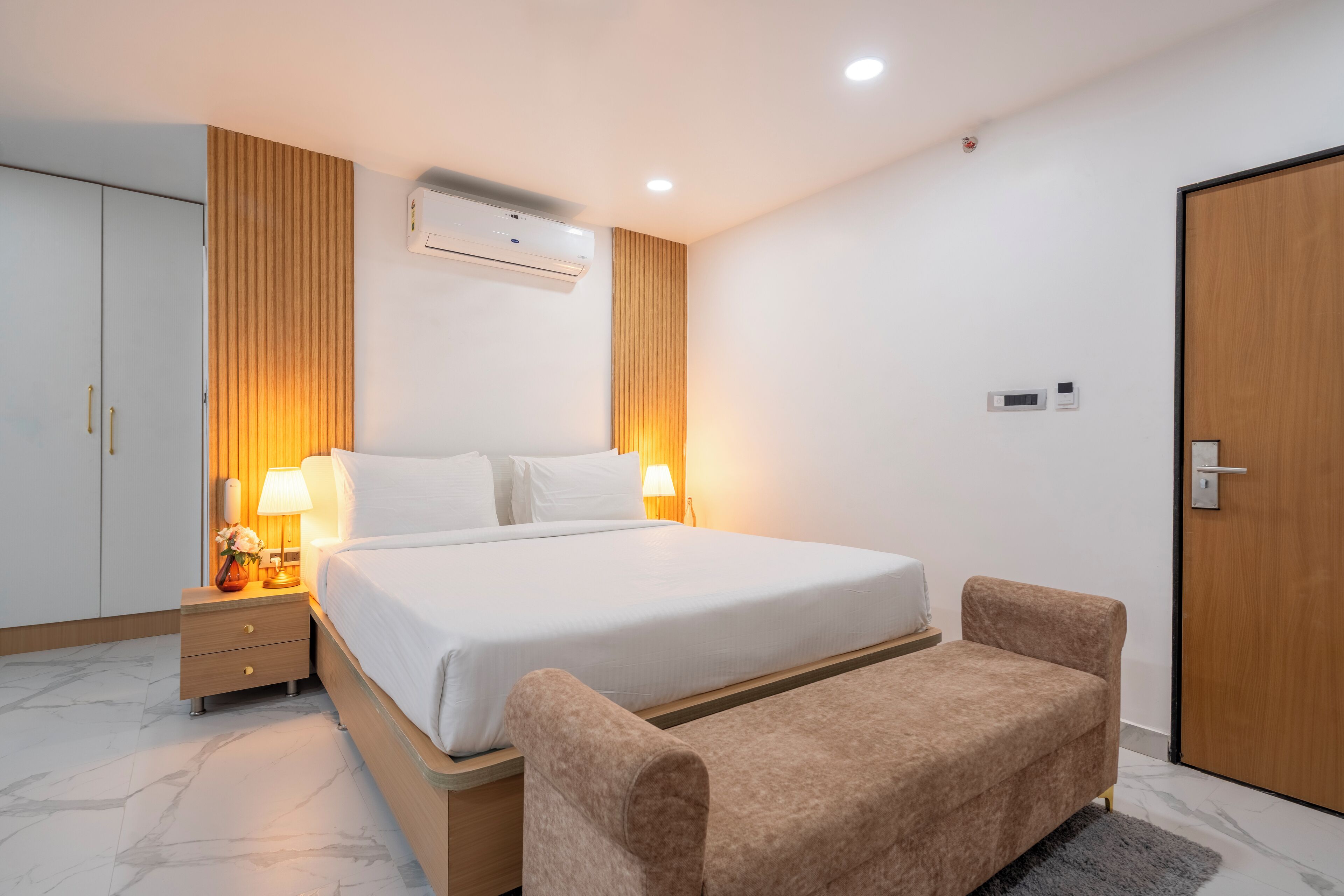 Quarto de hotel moderno com cama grande, luminárias laterais, guarda-roupa de madeira e um pequeno espaço de estar.