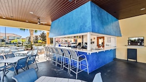 Uma área de bar ao ar livre com balcão azul, assentos com cadeiras de metal e vista para um bairro residencial com palmeiras.