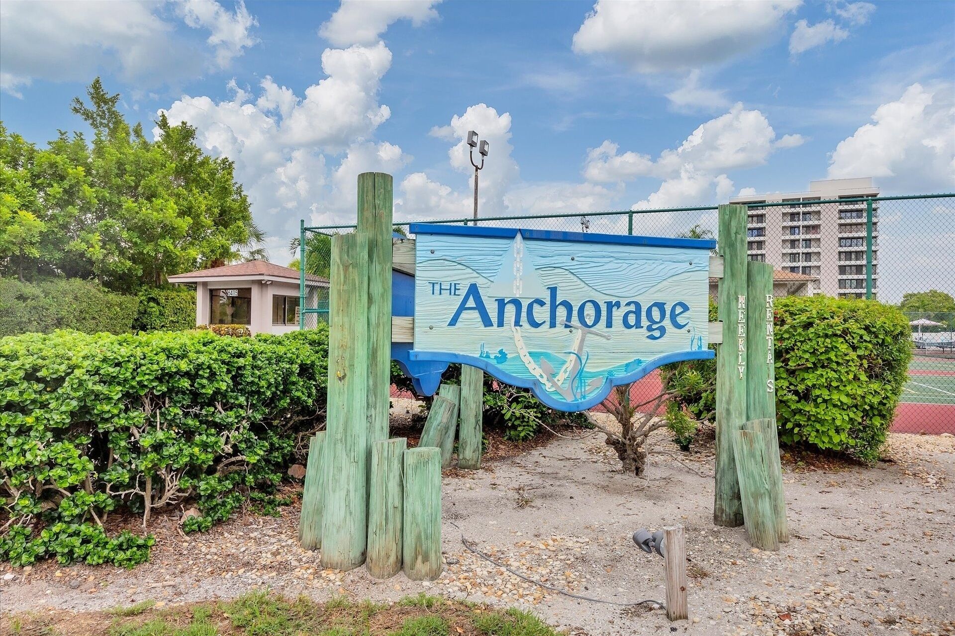 Placa para 'The Anchorage' com tema de praia, cercada por vegetação e um cercado.