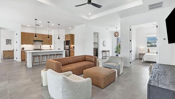 Ampla sala de estar com um grande sofá marrom, duas poltronas e uma mesa de centro. Cozinha com eletrodomésticos modernos e iluminação pendente visível ao fundo.
