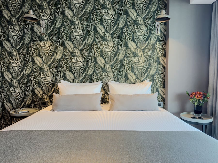 Uma cama bem arrumada com lençóis e almofadas brancas, papel de parede com padrão de folhas verdes e uma pequena mesa com um vaso de flores.