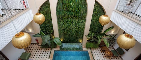 Uma área de piscina com uma piscina retangular central, rodeada por piso estampado, e ladeada por paredes de mosaico verde e lanternas penduradas.