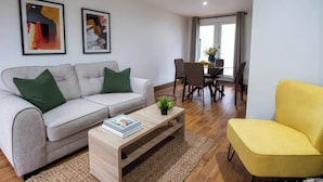 Uma sala de estar moderna com sofá, mesa de centro e poltrona amarela. Há duas pinturas abstratas emolduradas na parede, uma mesa de jantar com cadeiras e uma janela com cortinas.