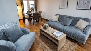 Uma sala de estar moderna com um sofá cinzento, uma poltrona a combinar e uma mesa de centro. É possível ver uma área de jantar com uma mesa e cadeiras através de uma porta.