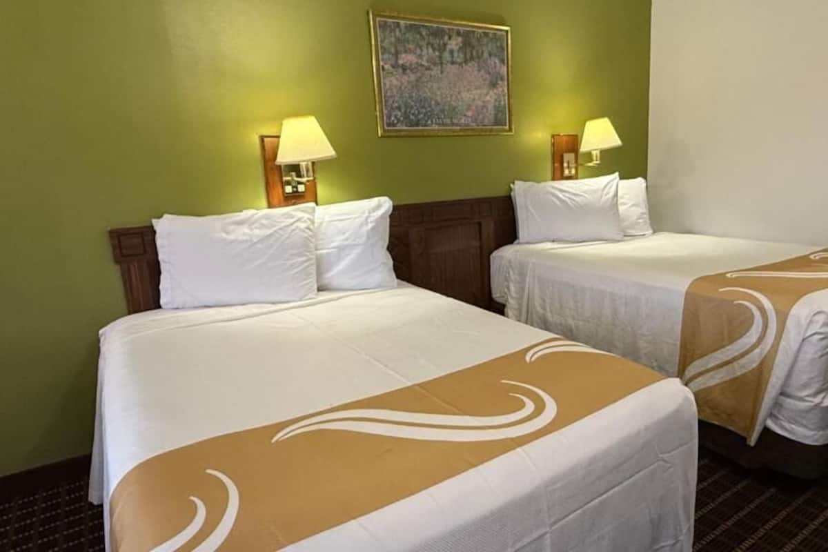 Duas camas de solteiro com lençóis brancos e detalhes decorativos marrons, uma parede verde e uma pintura acima das camas.