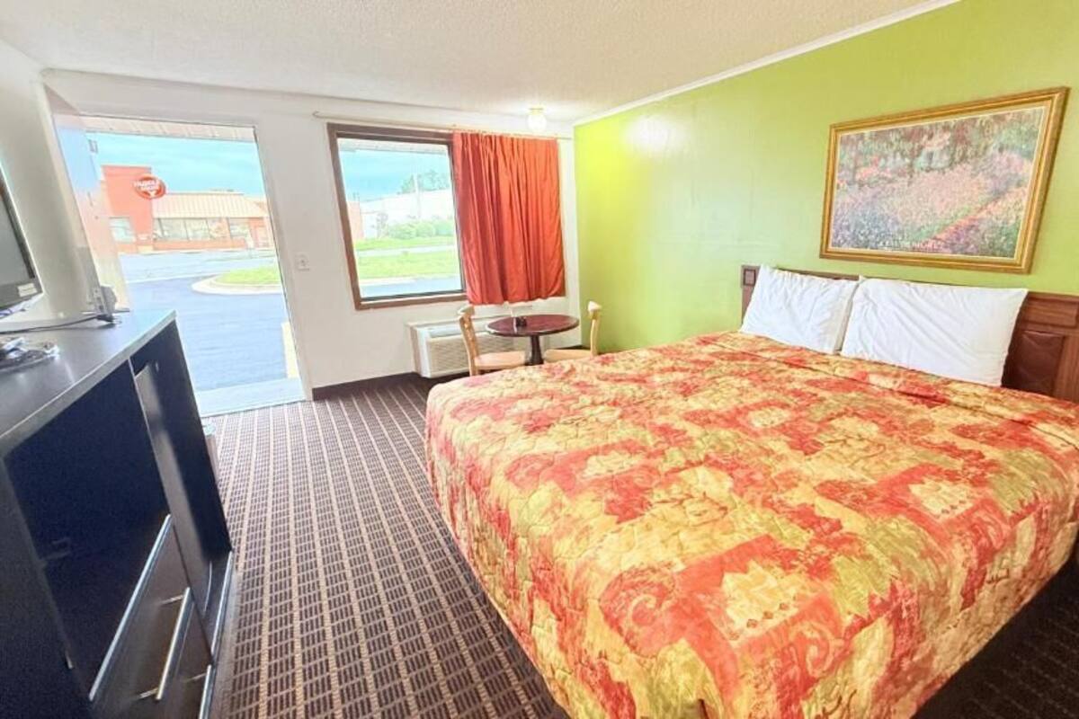 Quarto de hotel com cama, televisão, janela com cortinas e um quadro na parede.