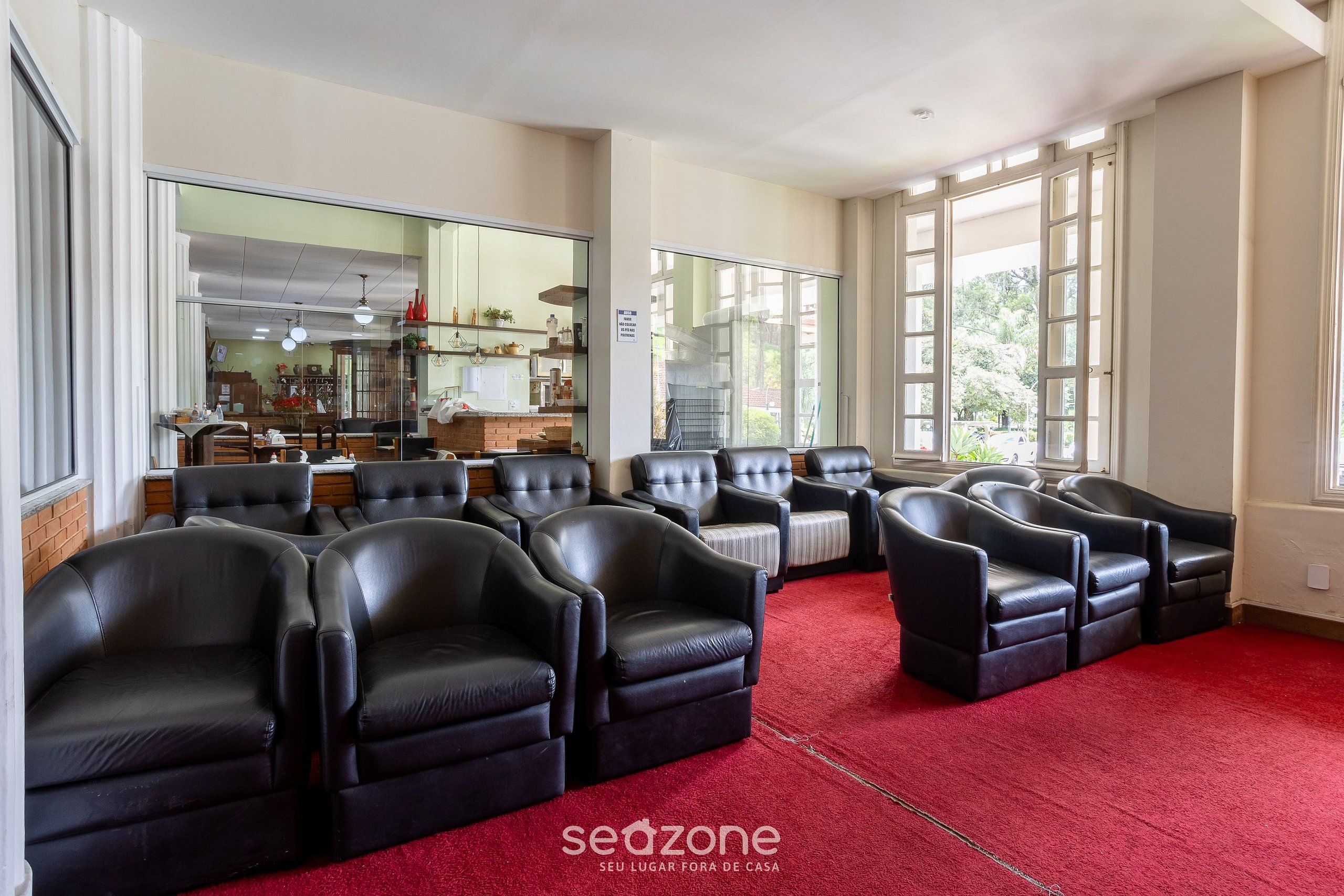 Quarto com poltronas de couro preto dispostas em semicírculo, tapete vermelho e grandes janelas com vista para o verde lá fora.