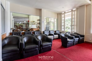 Quarto com poltronas de couro preto dispostas em semicírculo, tapete vermelho e grandes janelas com vista para o verde lá fora.