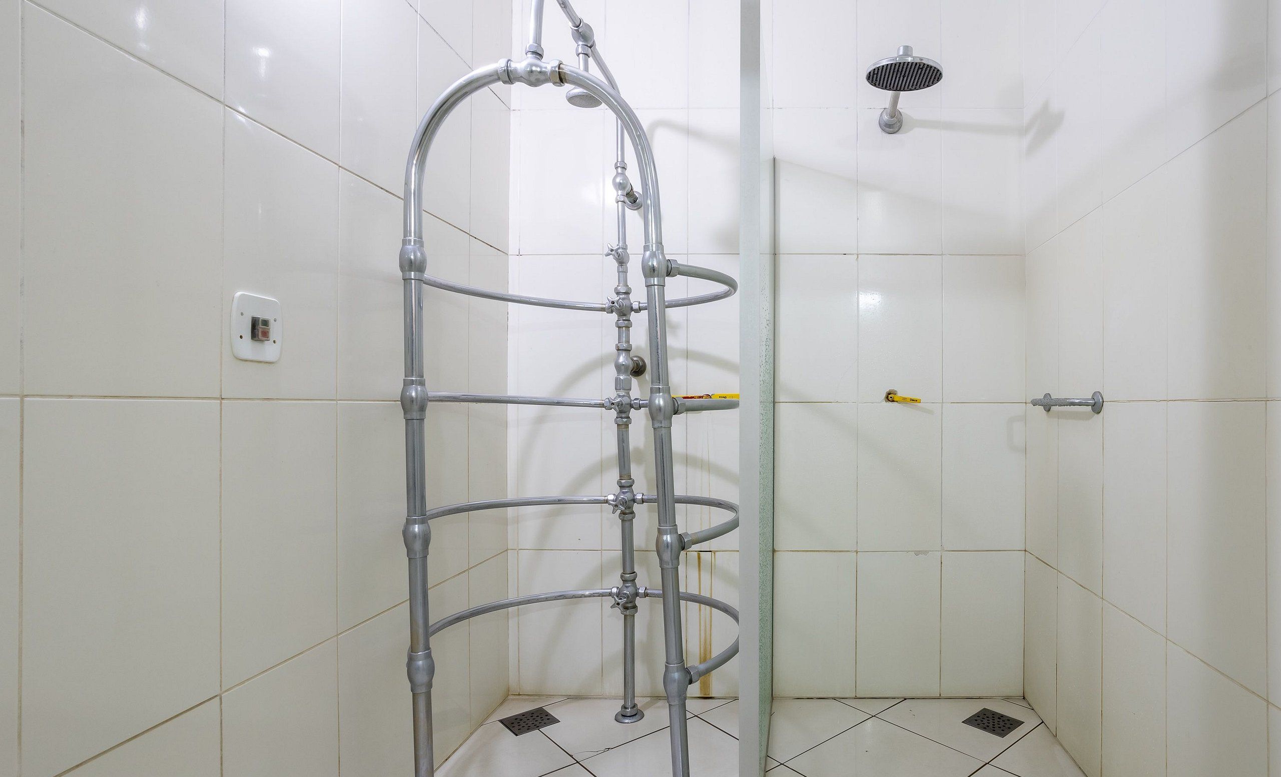 Chuveiro com estrutura metálica cromada, paredes revestidas de azulejo branco e ralo no chão.