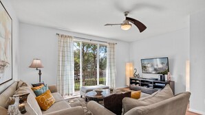 Uma sala de estar com sofá, poltronas, mesa de centro, televisão e ventoinha no teto.