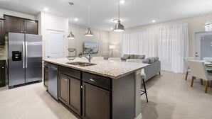 Cozinha moderna com eletrodomésticos de aço inoxidável, ilha com bancada de granito, iluminação pendente e área de jantar com sofá e cadeiras.