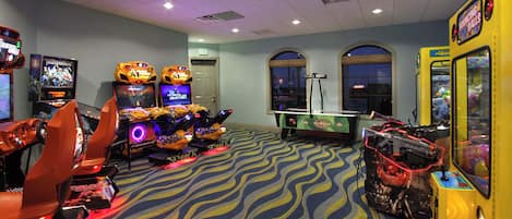 Uma área de jogos de fliperama com jogos de corrida e de garra, mesa de bilhar e vista para o exterior através de janelas em arco.