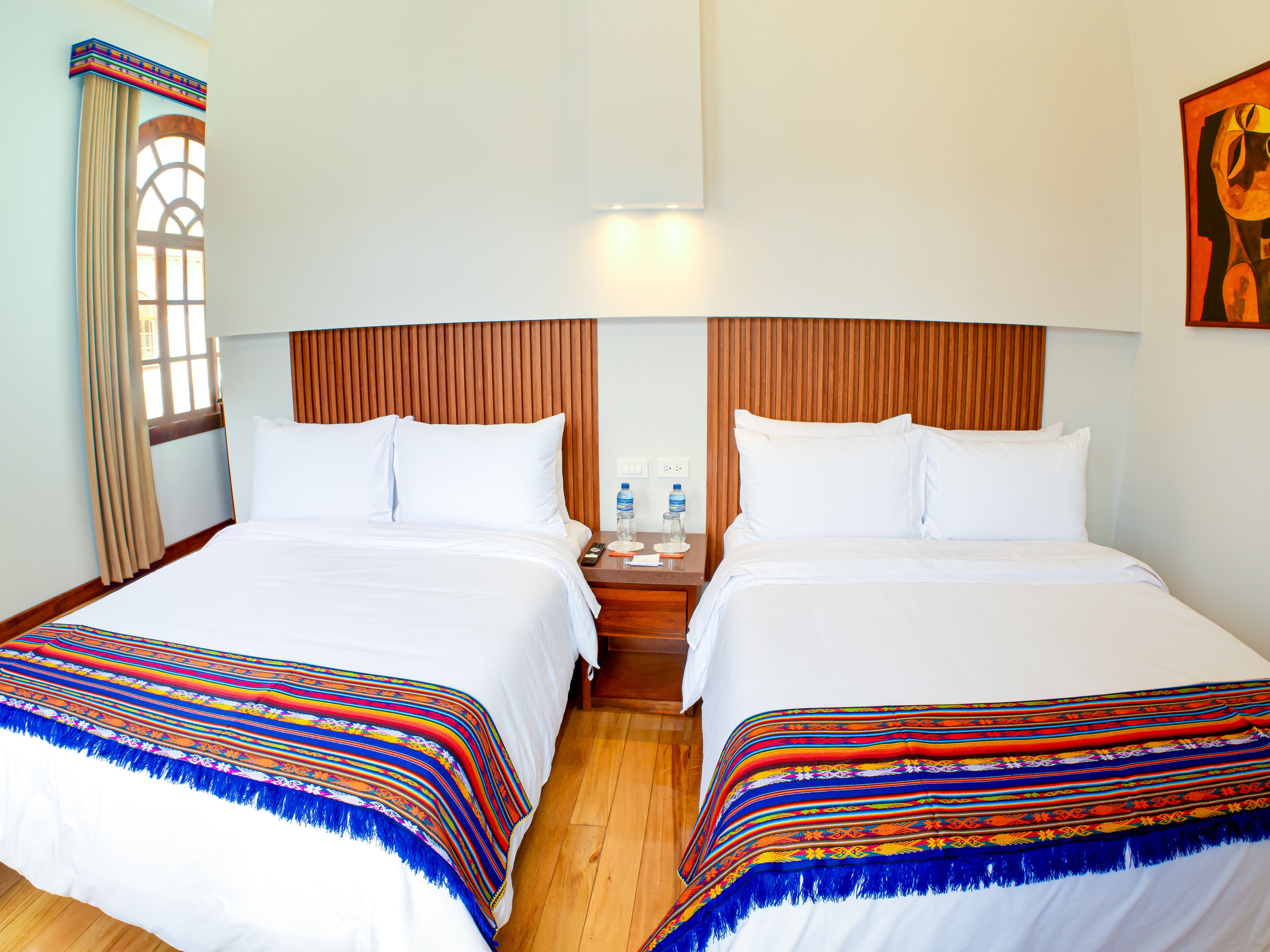 Quarto de hotel com duas camas, cabeceiras de madeira, mesa de cabeceira com luminária e janela com cortinas.