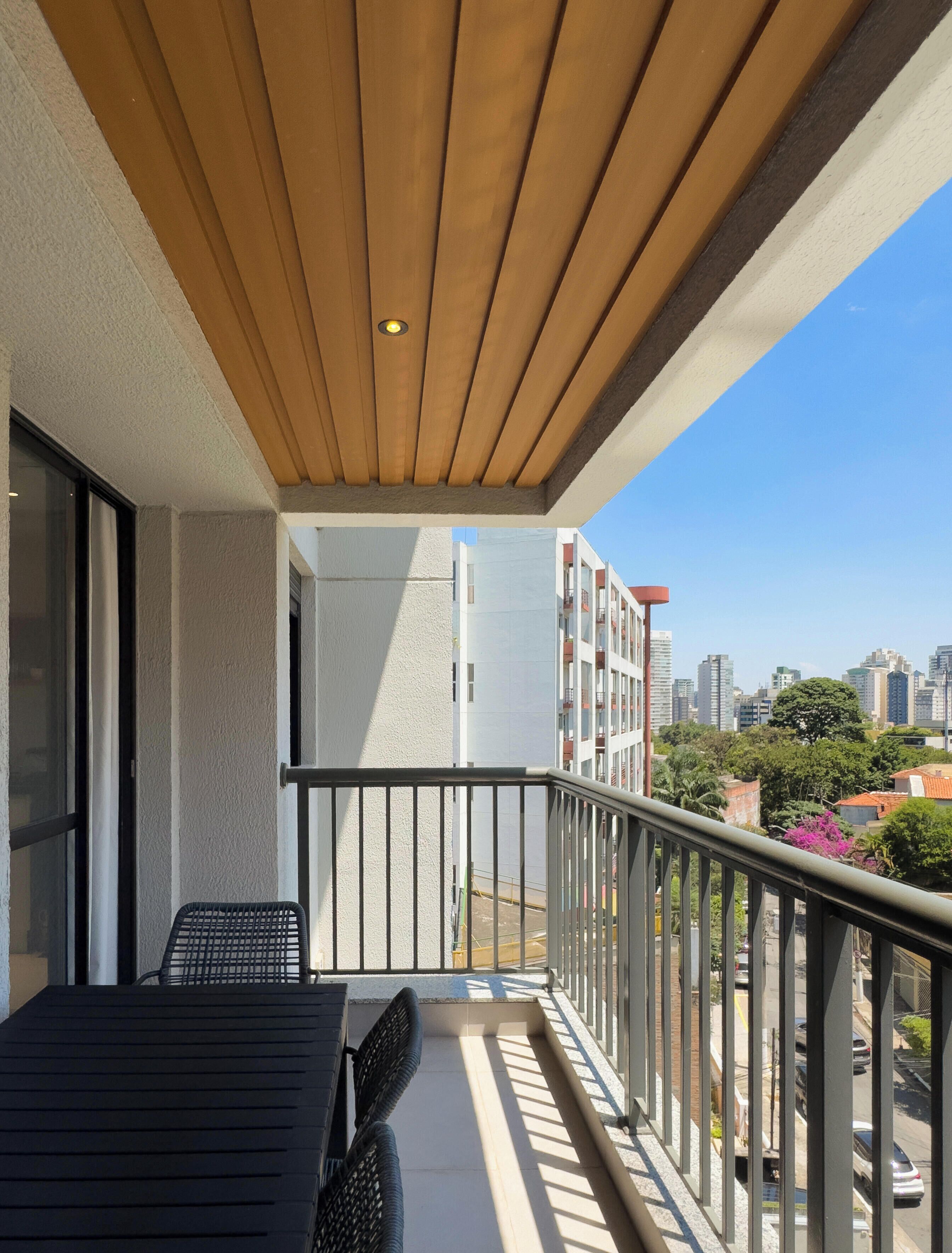 Uma varanda com teto de madeira, guarda-corpo de metal e vista para a paisagem urbana.