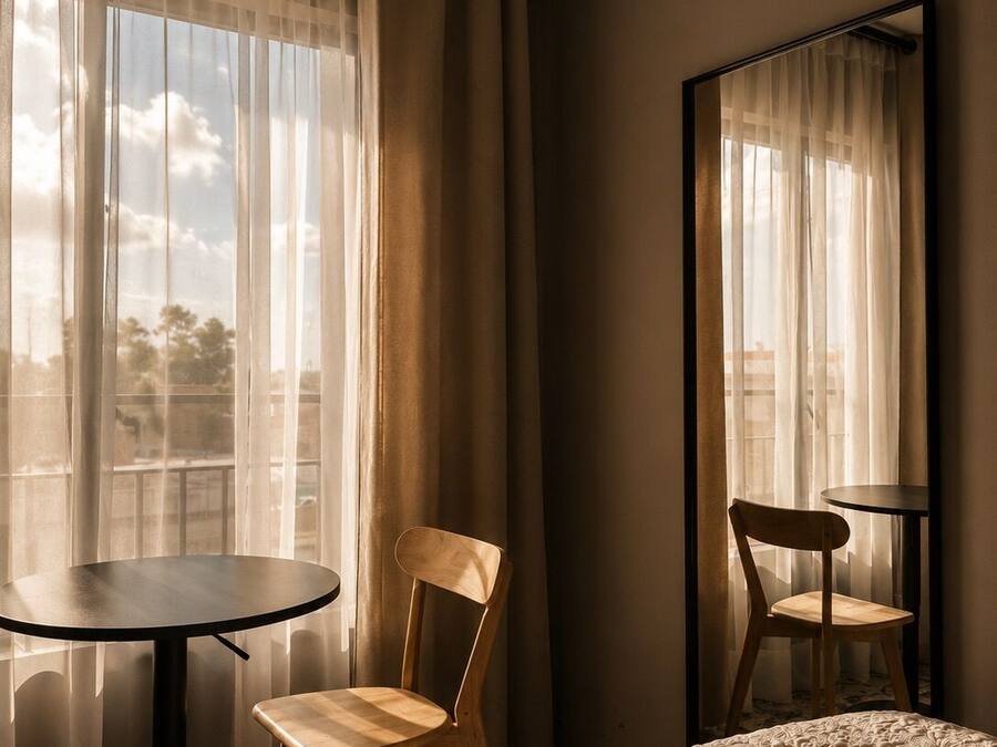 Quarto de hotel com cama, duas cadeiras, mesa redonda, espelho grande e cortinas leves.