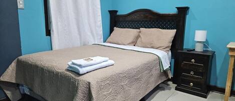 Cama bem arrumada com colcha estampada e travesseiros, mesa de cabeceira com abajur e criado-mudo com gaveta.