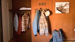 Quarto com parede de madeira, um cabideiro com vários casacos, uma cama com travesseiro branco e uma imagem de cabeça de veado montada.