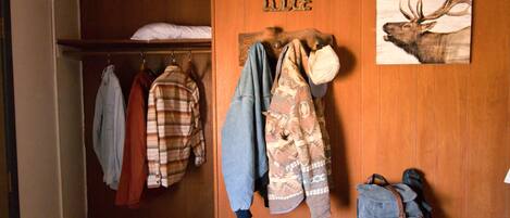 Quarto com parede de madeira, um cabideiro com vários casacos, uma cama com travesseiro branco e uma imagem de cabeça de veado montada.