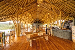 Uma área de refeições com telhado de bambu, mesas e cadeiras de madeira, um bar e vista para a paisagem.