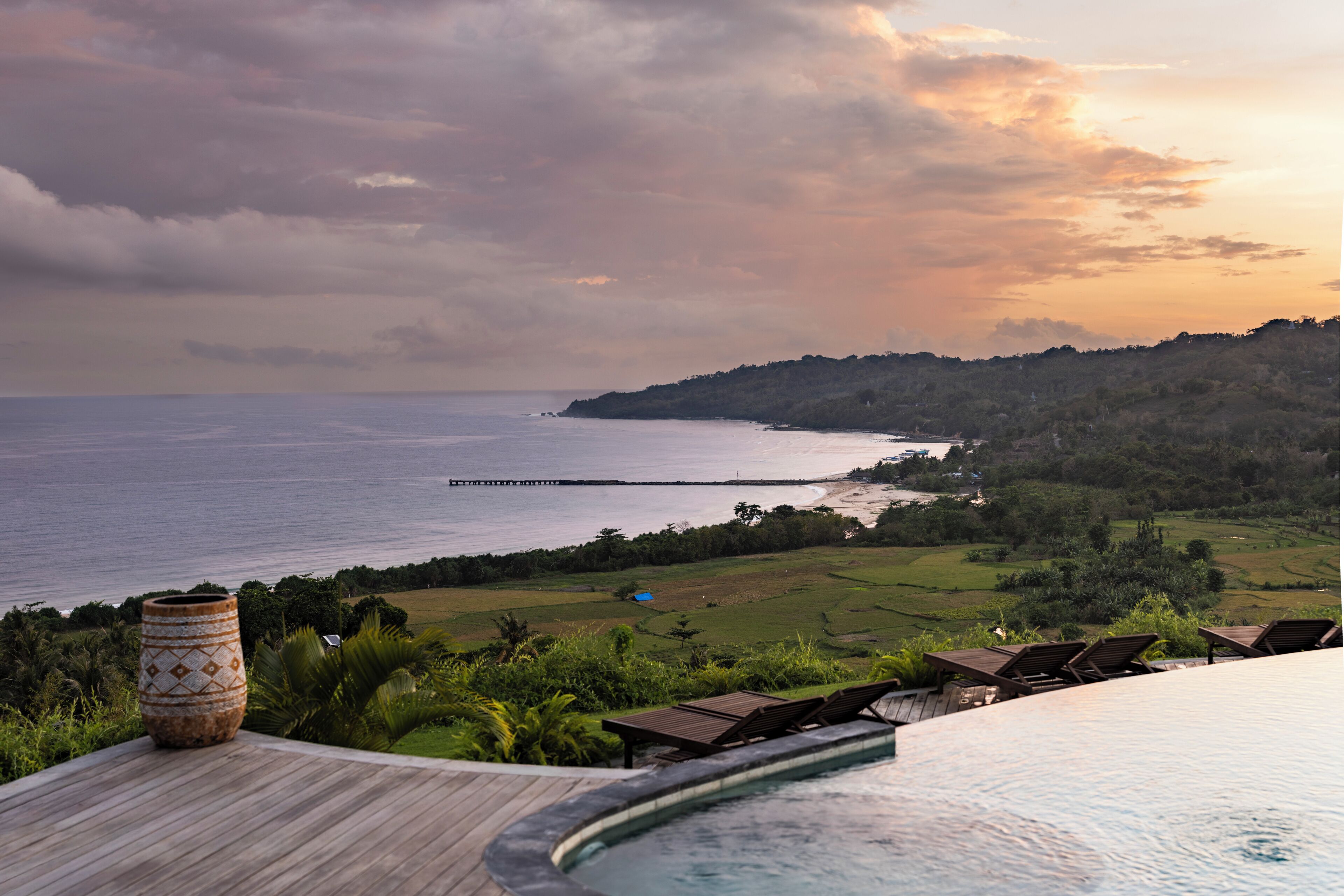Área de piscina com vista para o oceano e um píer, rodeada por vegetação e espreguiçadeiras.