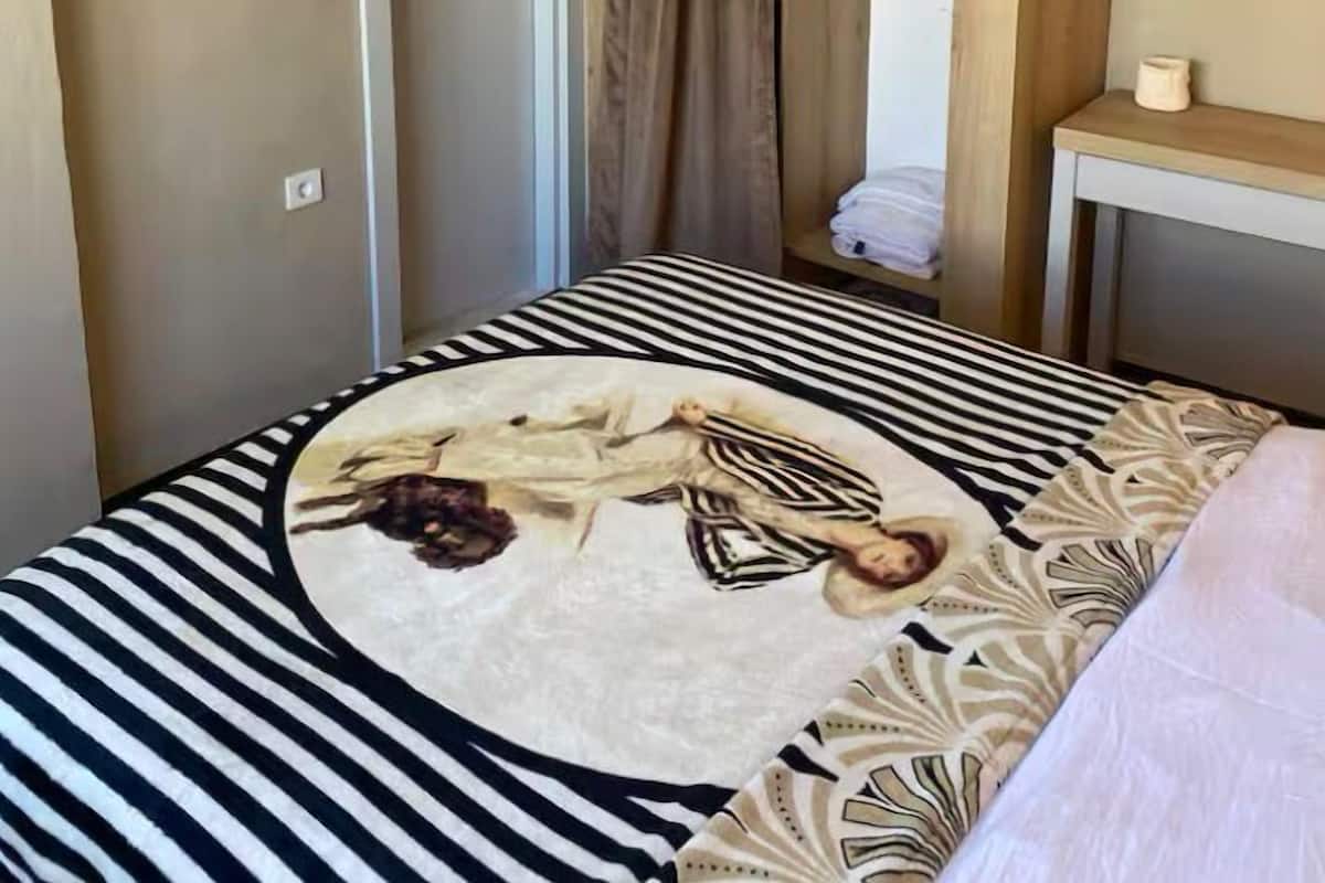 Quarto de hotel com cama coberta por um edredom listrado, luminária pendente, guarda-roupa e uma pequena mesa de cabeceira.