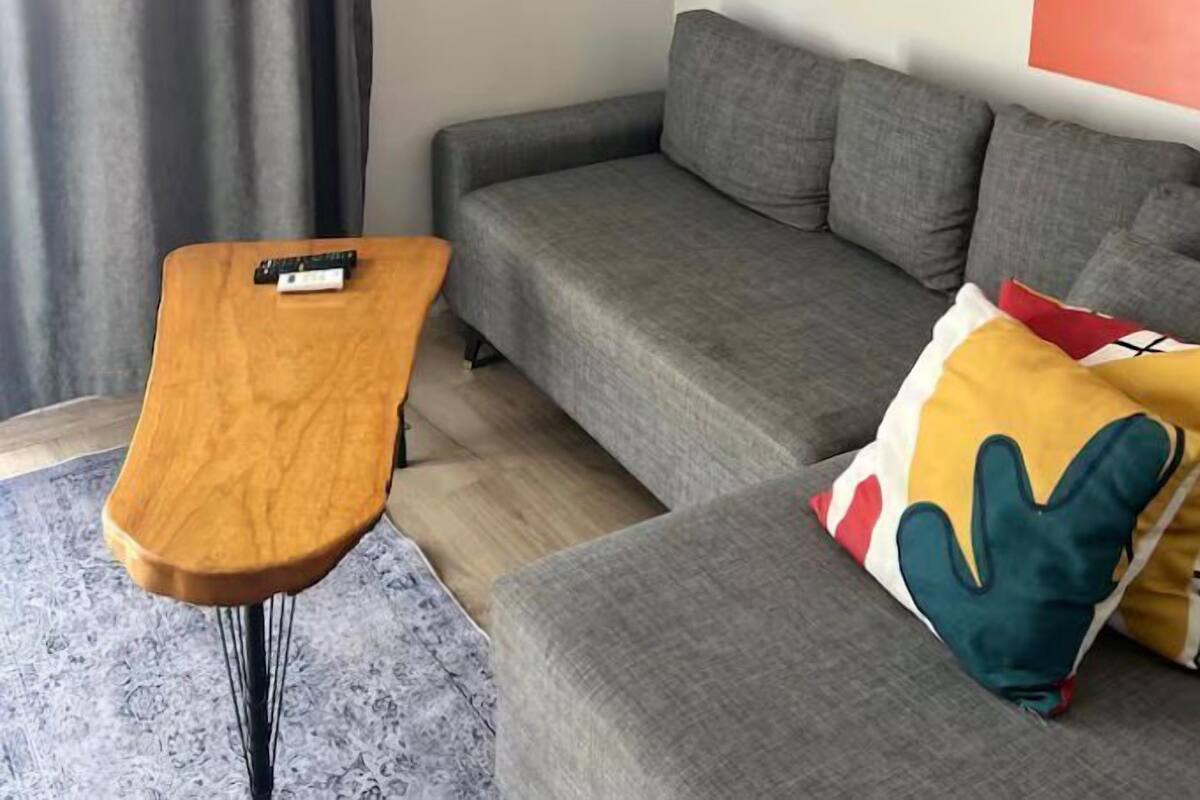Uma sala de estar moderna com um sofá cinzento, uma mesa lateral de madeira e uma obra de arte colorida na parede.