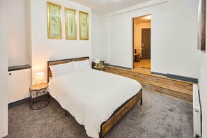 Quarto com cama de madeira, mesinha de cabeceira com luminária e três quadros de plantas na parede.