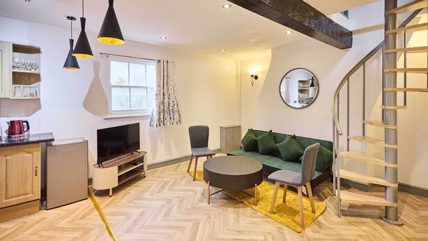 Uma sala de estar moderna com piso em padrão herringbone, um conjunto de sofá, uma mesa de centro e uma televisão. Há uma área de cozinha com armários e uma geladeira. Uma escada em espiral é visível ao fundo.