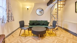 Uma sala de estar moderna com um sofá verde, duas cadeiras cinzentas, uma mesa de centro redonda e um chão de madeira em padrão herringbone.