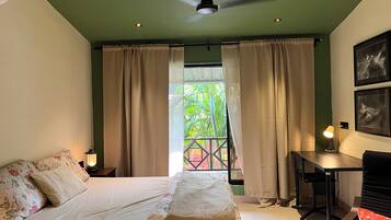 Quarto com cama, escrivaninha, cadeira, ventilador de teto e janela com cortinas.