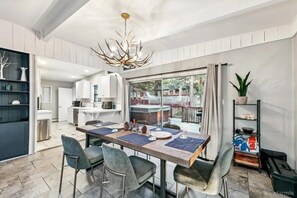 Uma sala de jantar com uma mesa grande posta para quatro pessoas, cercada por cadeiras. Há um lustre sobre a mesa e uma porta de vidro deslizante que dá para um pátio externo.