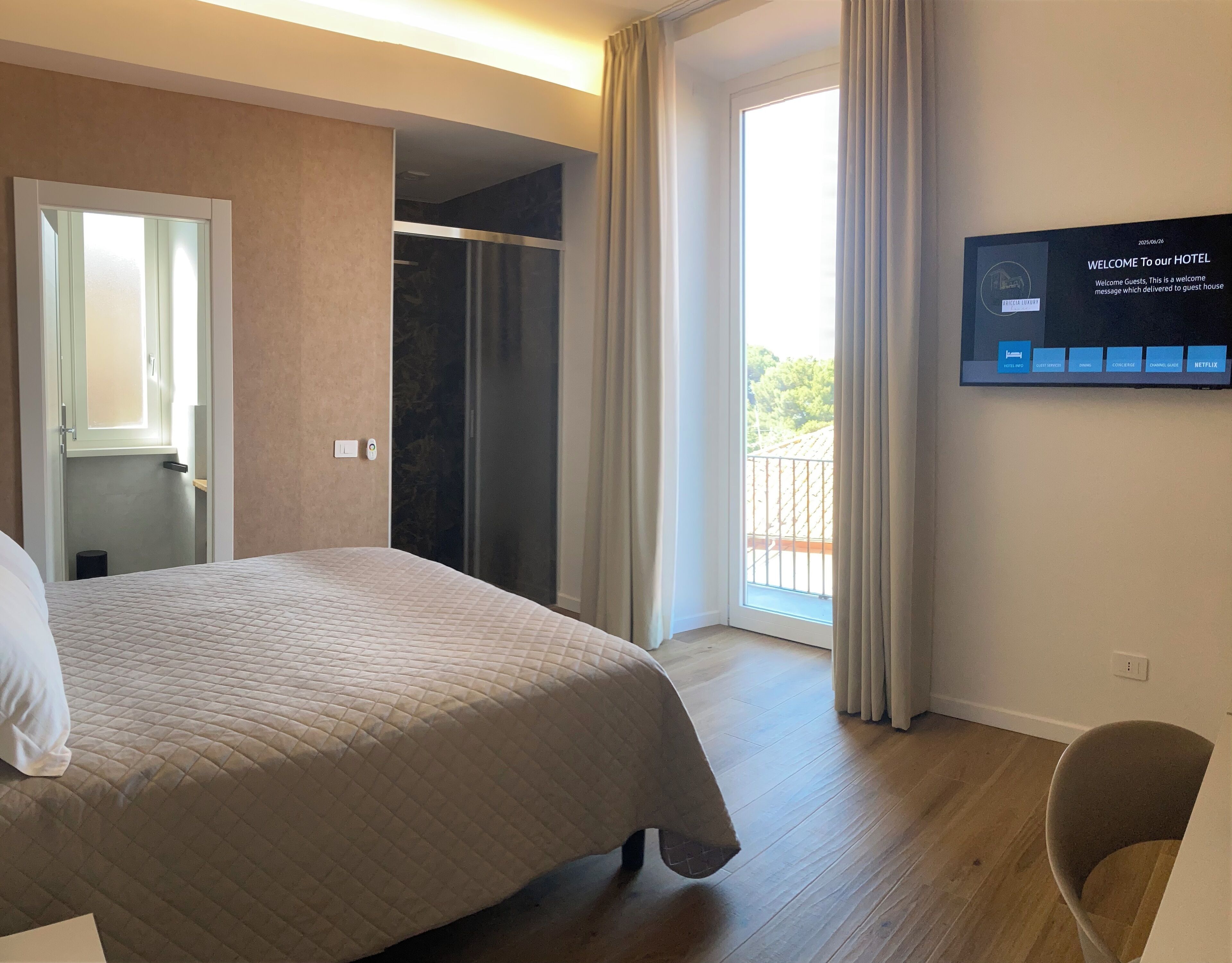 Quarto de hotel com uma cama, televisão fixada na parede, porta de correr de vidro para a varanda e área de chuveiro.