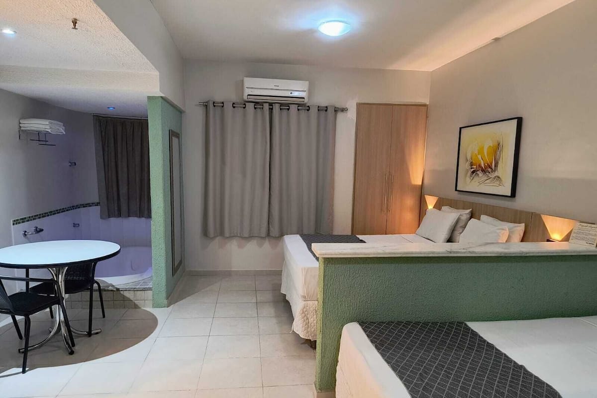 Quarto de hotel com cama, uma pequena mesa com cadeiras, ar condicionado e um quadro na parede.