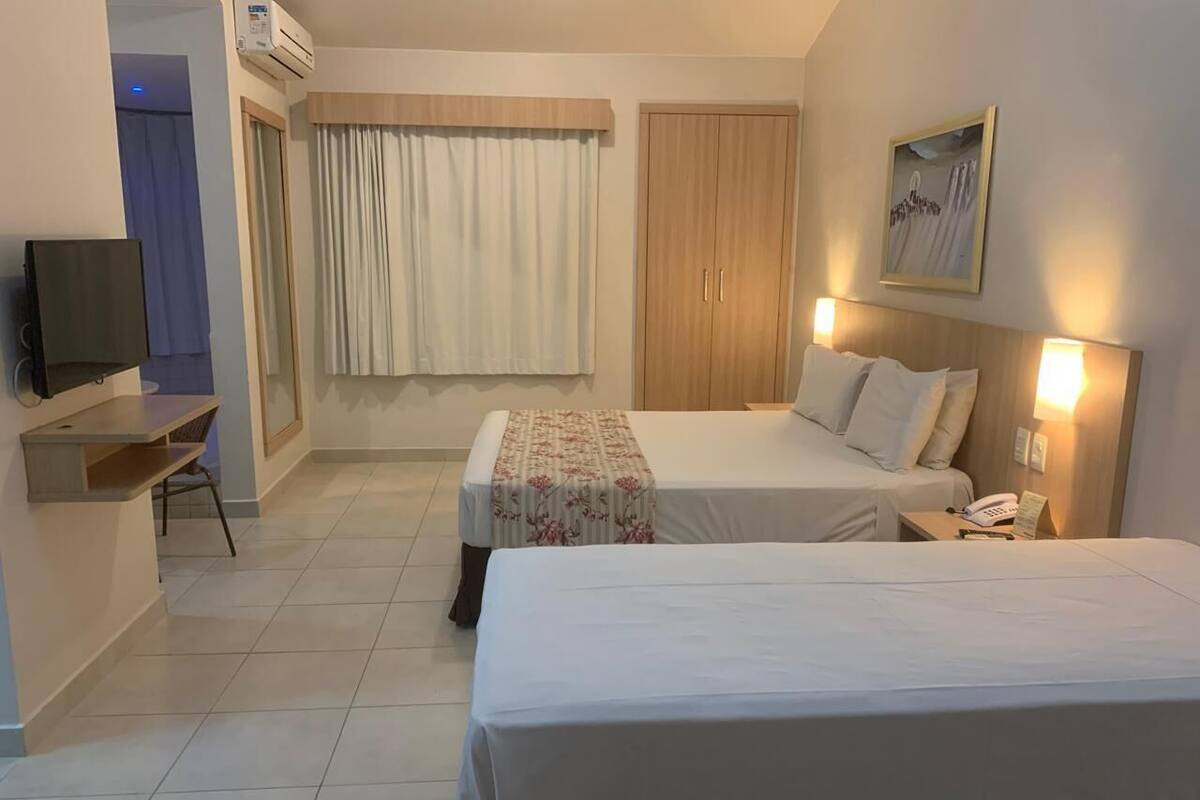 Quarto de hotel com duas camas, uma escrivaninha com cadeira, televisão e guarda-roupa.