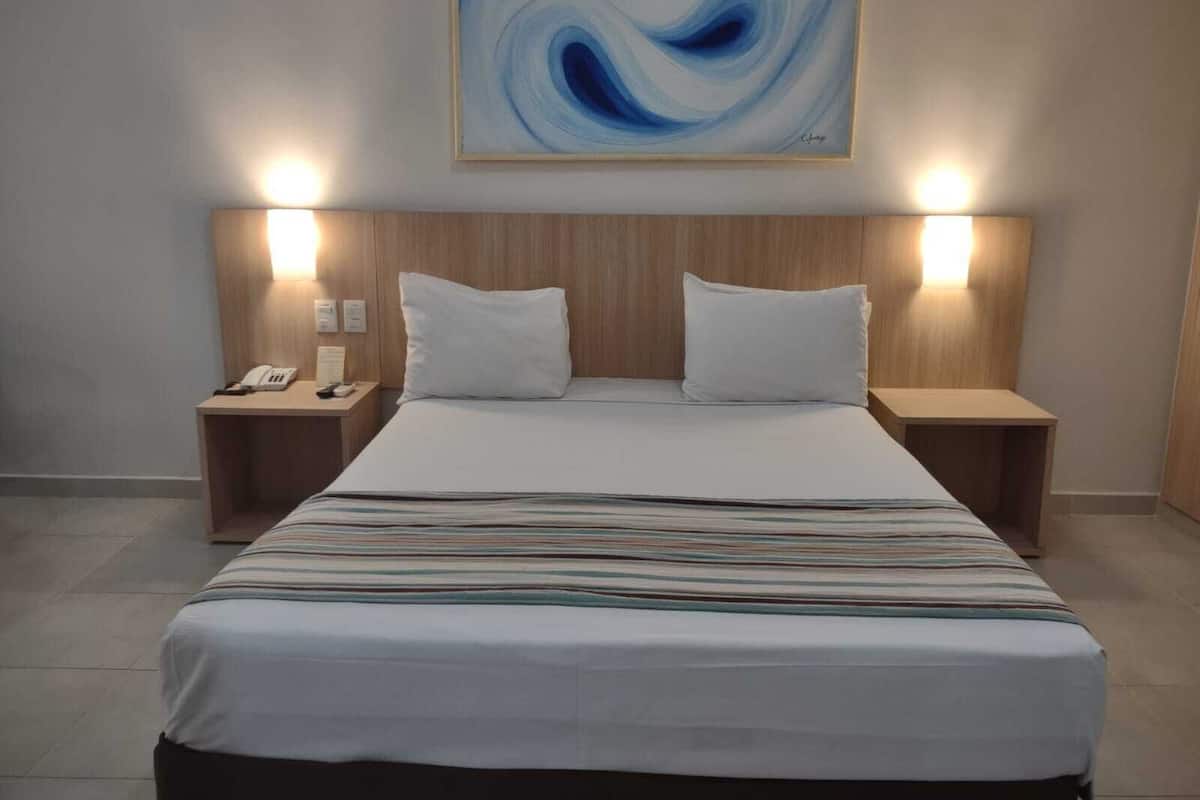 Quarto de hotel com uma cama grande, duas mesas de cabeceira, um telefone e um quadro na parede.