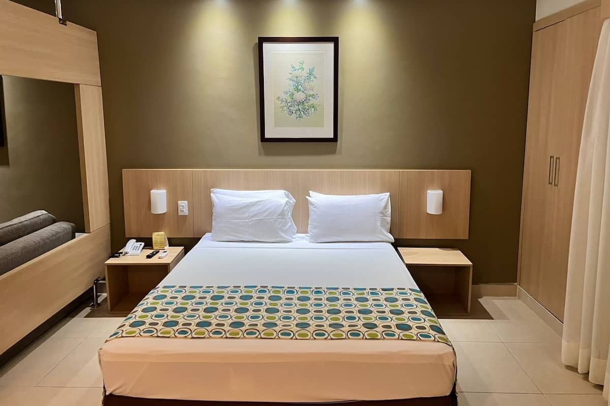 Quarto de hotel com uma cama grande, duas mesas de cabeceira, um telefone fixado na parede e uma obra de arte emoldurada acima da cama.