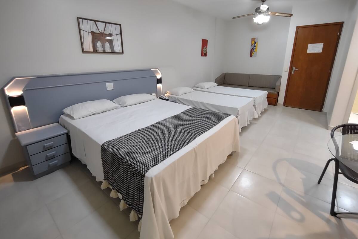 Quarto de hotel com uma cama grande, duas camas menores, uma escrivaninha e uma cadeira.