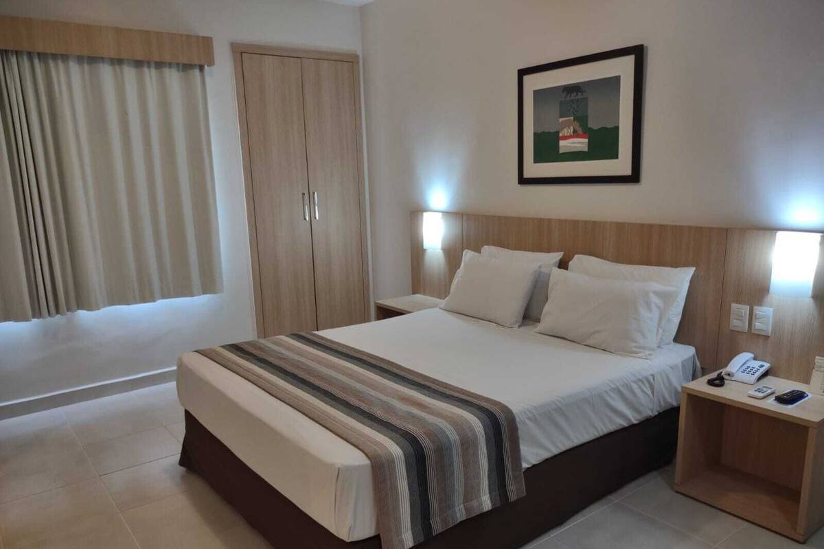 Quarto de hotel com uma cama grande, um guarda-roupa de madeira, uma mesa de cabeceira com um telefone e um quadro na parede.