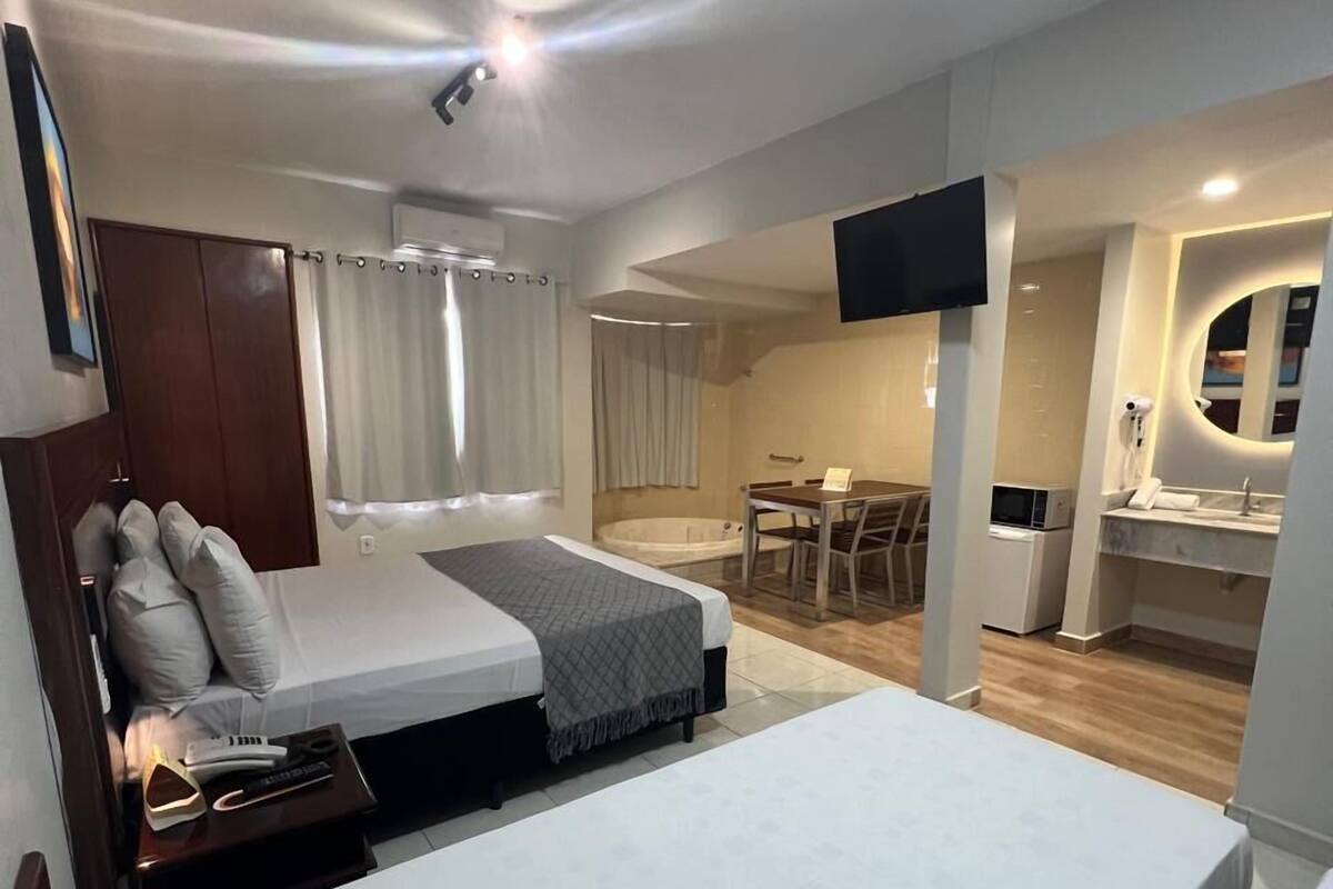 Quarto de hotel moderno com uma cama grande, uma escrivaninha, uma cadeira, uma televisão e um banheiro com espelho e pia.