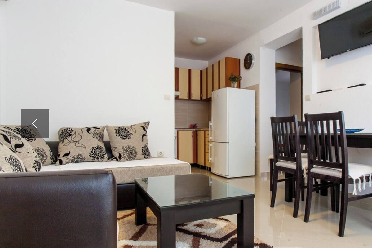 Sala de estar com sofá, mesa de centro de vidro e área de jantar com mesa e cadeiras. Uma TV está fixada na parede acima da área de jantar.