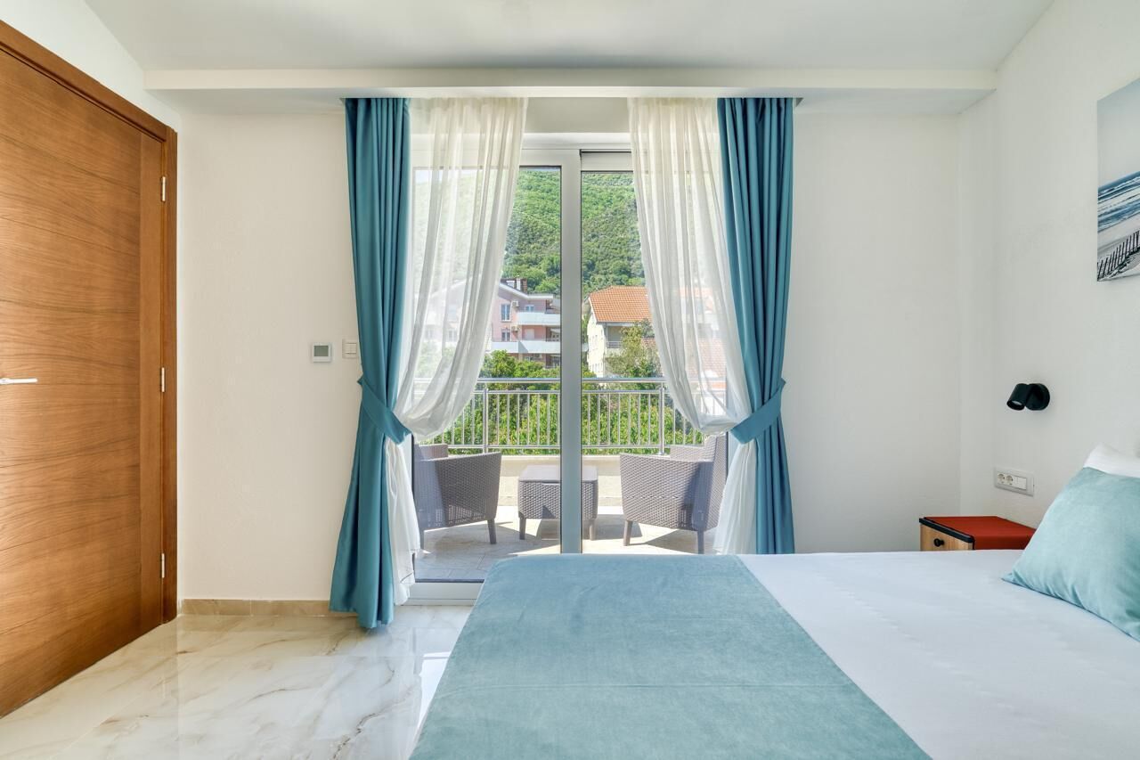 Quarto com cama, guarda-roupa de madeira, varanda com mesa e cadeiras e vista para edifícios.