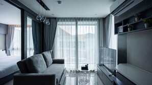 Quarto de hotel moderno com sofá, mesinha redonda, televisão e janela grande com cortinas leves.