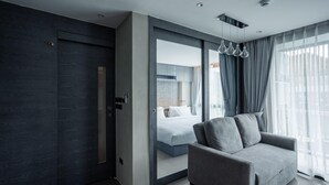 Quarto de hotel moderno com sofá cinzento, cama com roupa de cama branca, janela grande com cortinas leves e porta de madeira escura.